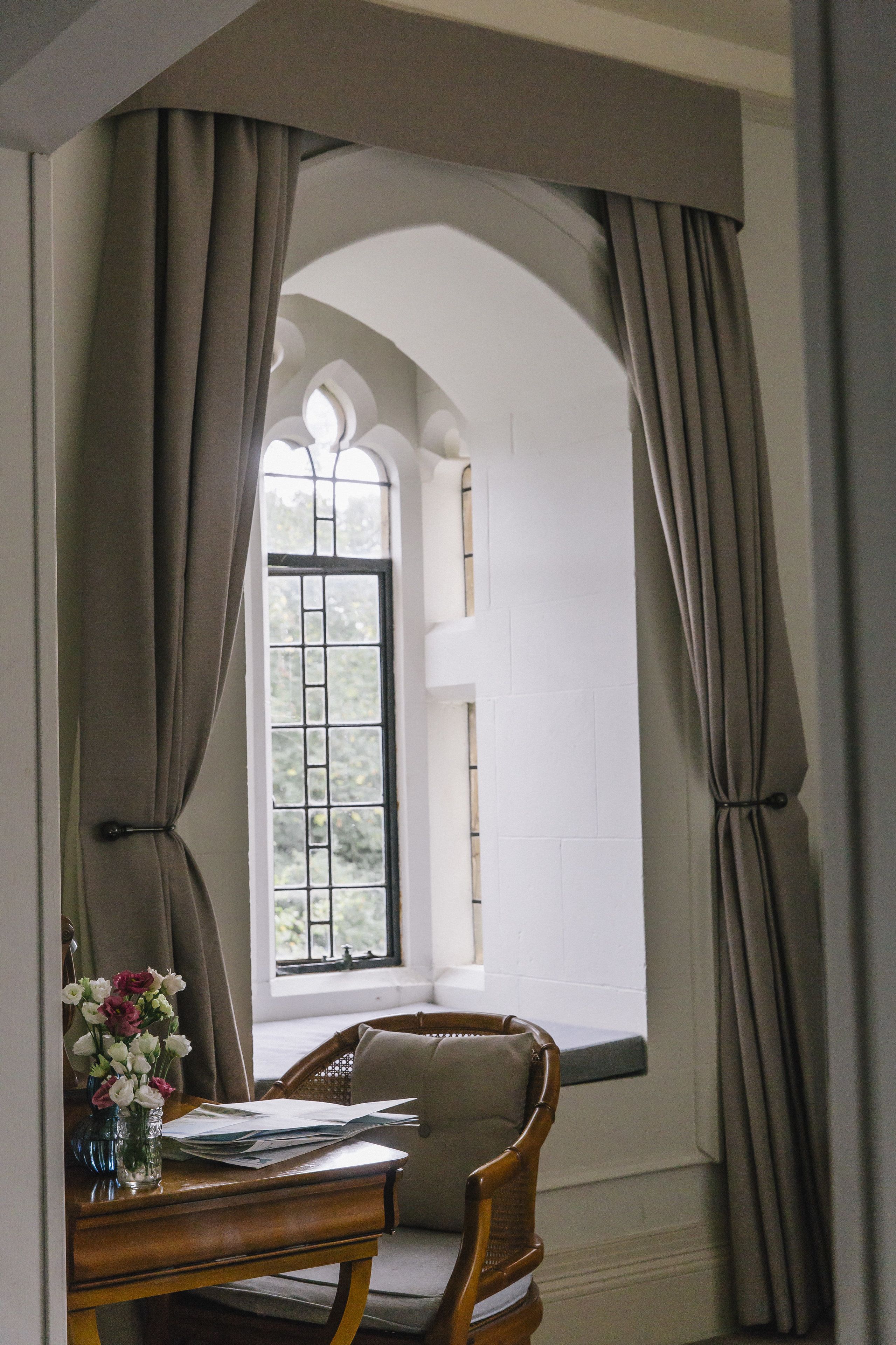 Quarto com janela, cortinas, uma cadeira e uma mesa com papéis e um vaso de flores.