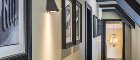 Um corredor com obras de arte emolduradas nas paredes, uma escada com grades pretas e um lustre ao fundo.
