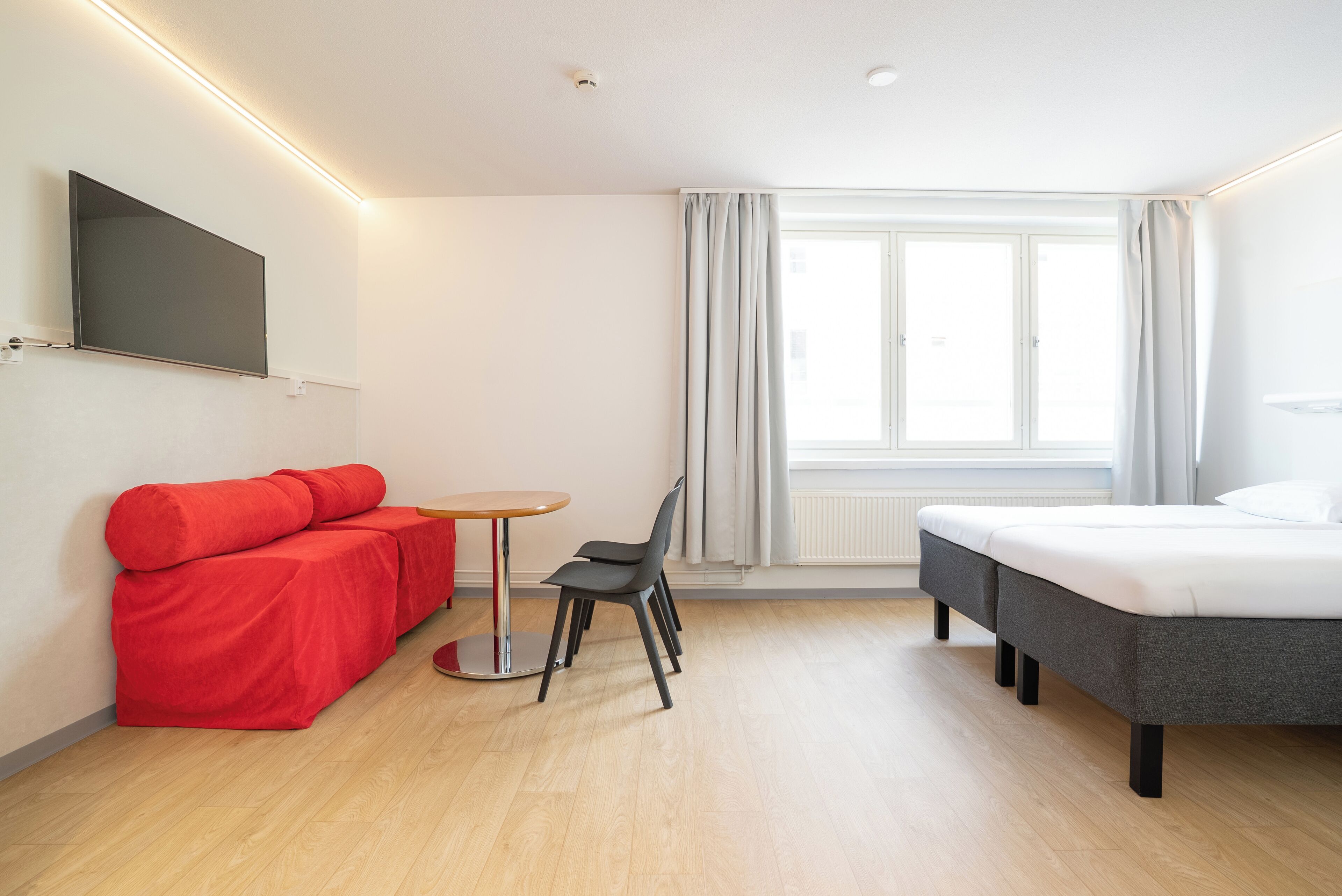 Quarto de hotel moderno com uma cama, um sofá vermelho, uma pequena mesa redonda e uma TV fixada na parede.