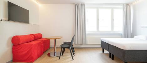 Quarto de hotel moderno com uma cama, um sofá vermelho, uma pequena mesa redonda e uma TV fixada na parede.