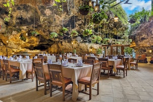 Um restaurante com mesas preparadas para refeições, um interior semelhante a uma caverna com plantas penduradas e um paredão rochoso ao fundo.