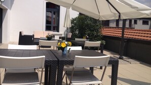 Um pátio ao ar livre com mesa, cadeiras, um guarda-sol grande e plantas em vasos.