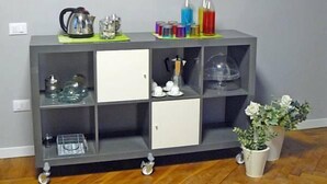 Um armário cinzento com vários compartimentos, alguns com portas de vidro, e um pequeno vaso com planta no chão.