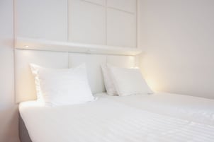 Cama com roupa de cama e almofadas brancas, cabeceira com luminária, e parede lisa.