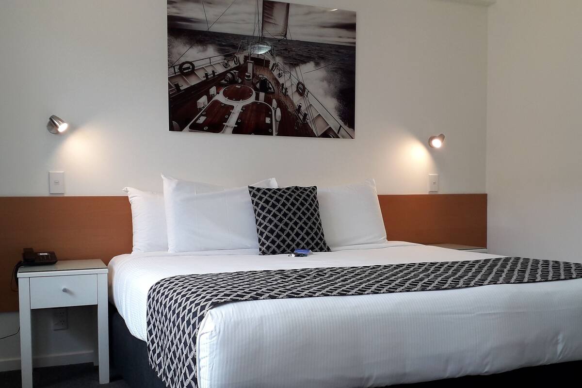 Quarto de hotel com uma cama grande, uma mesa de cabeceira, um quadro pendurado na parede e duas luminárias de parede.