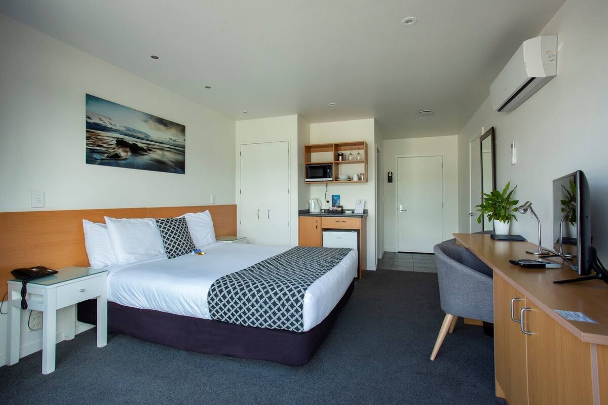 Quarto de hotel com cama, mesa de cabeceira, escrivaninha e cozinha compacta.