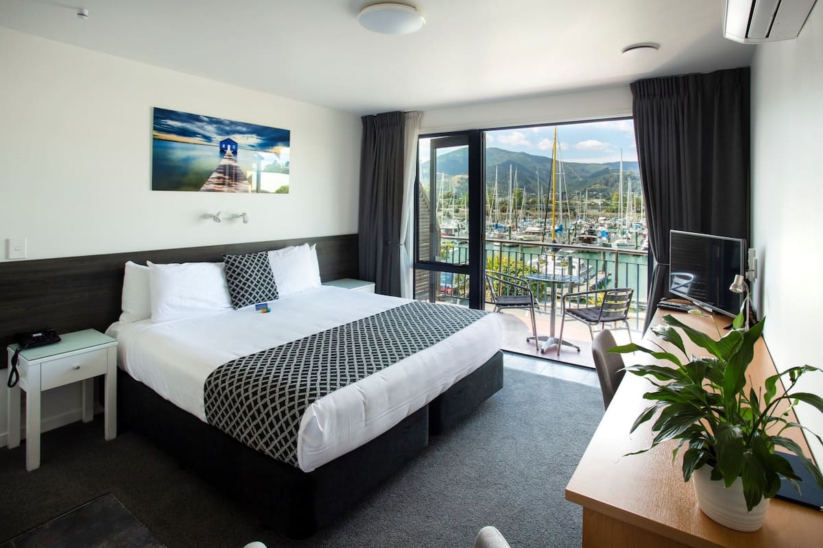 Quarto de hotel com uma cama grande, uma escrivaninha com computador, uma planta em vaso e uma varanda com vista para barcos e montanhas.