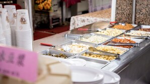 Buffet com diversos alimentos em bandejas metálicas, pratos empilhados à esquerda e uma placa rosa em primeiro plano.