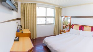 Quarto de hotel com cama, mesa de cabeceira, escrivaninha com telefone, espelho e janela com cortinas.