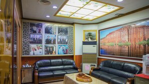 Um hall com dois sofás de couro preto, uma mesa de centro de madeira, piso ladrilhado, parede com fotos emolduradas, uma pintura grande e teto com claraboia.
