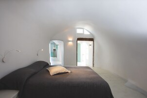 Quarto com uma cama de cor escura, um travesseiro claro e uma pequena mesa de cabeceira. Há uma janela com vista para um corredor e uma porta.