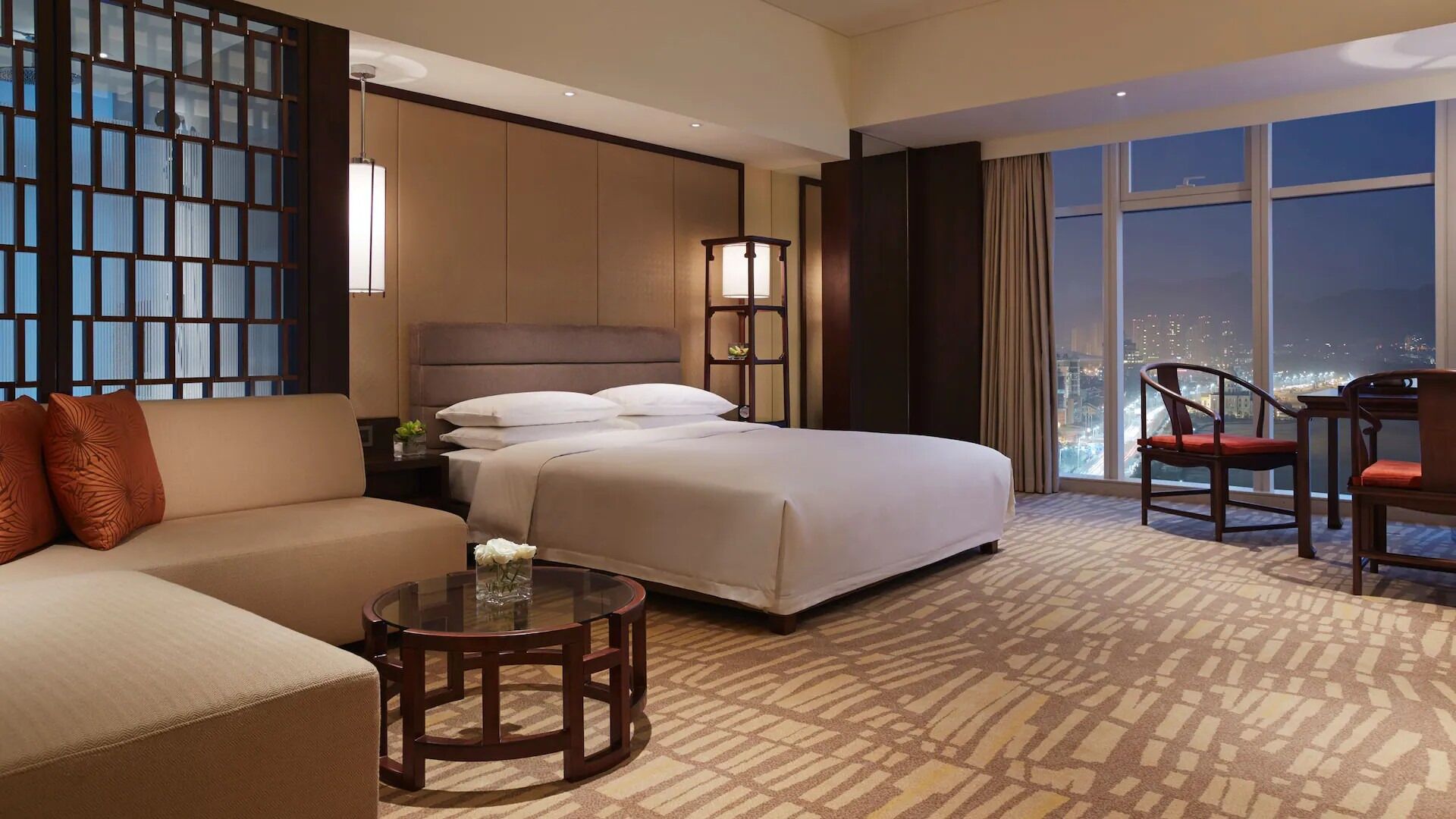 Quarto de hotel com cama, sofá, uma mesa pequena e vista da cidade à noite.