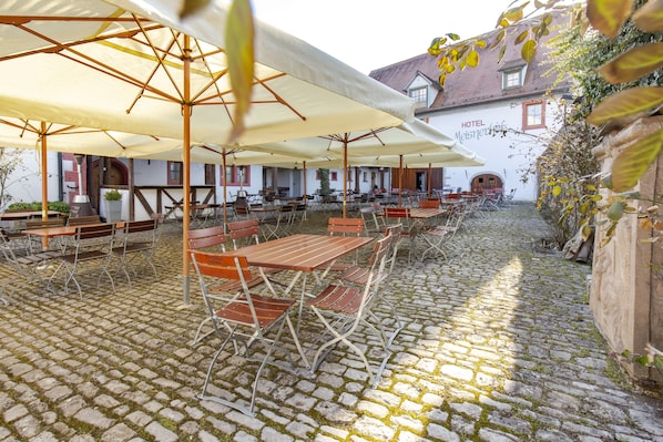 Um café ao ar livre com mesas e cadeiras sob guarda-sóis, um caminho de paralelepípedos e um edifício com a inscrição "HOTEL Meissnerstrasse".