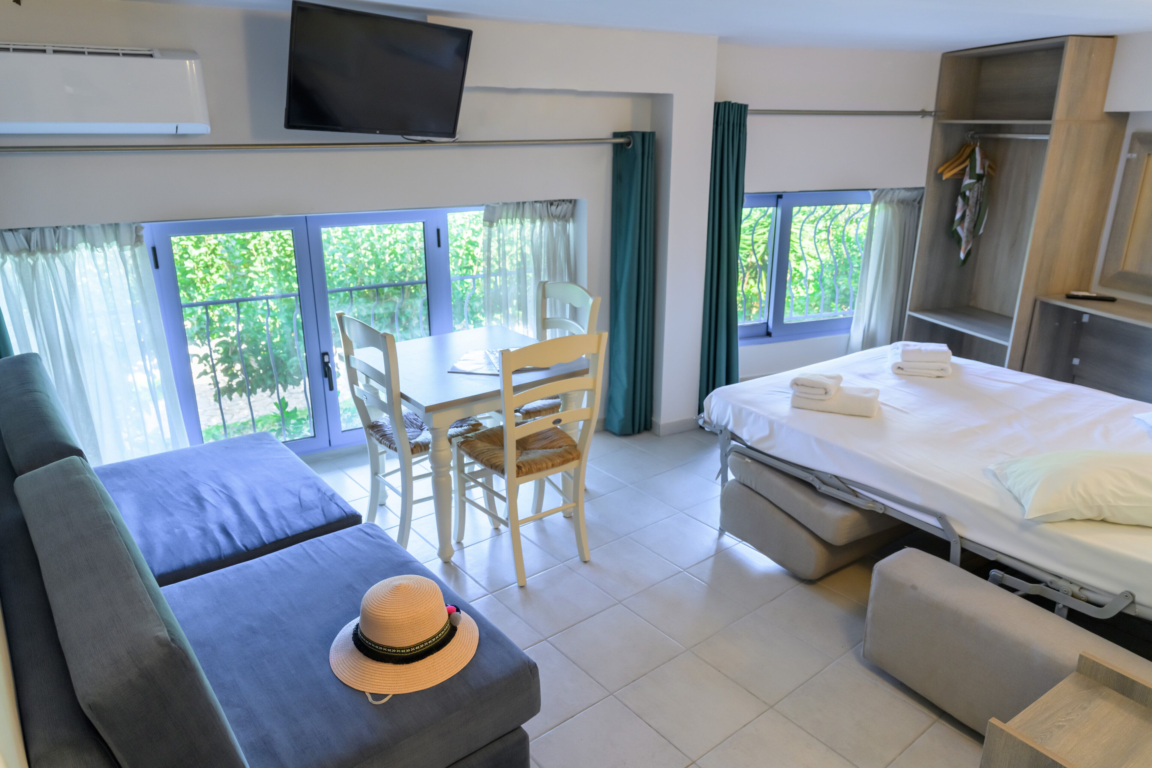 Quarto de hotel com cama, sofá, mesa de jantar com cadeiras, TV fixada na parede e grandes janelas com cortinas.