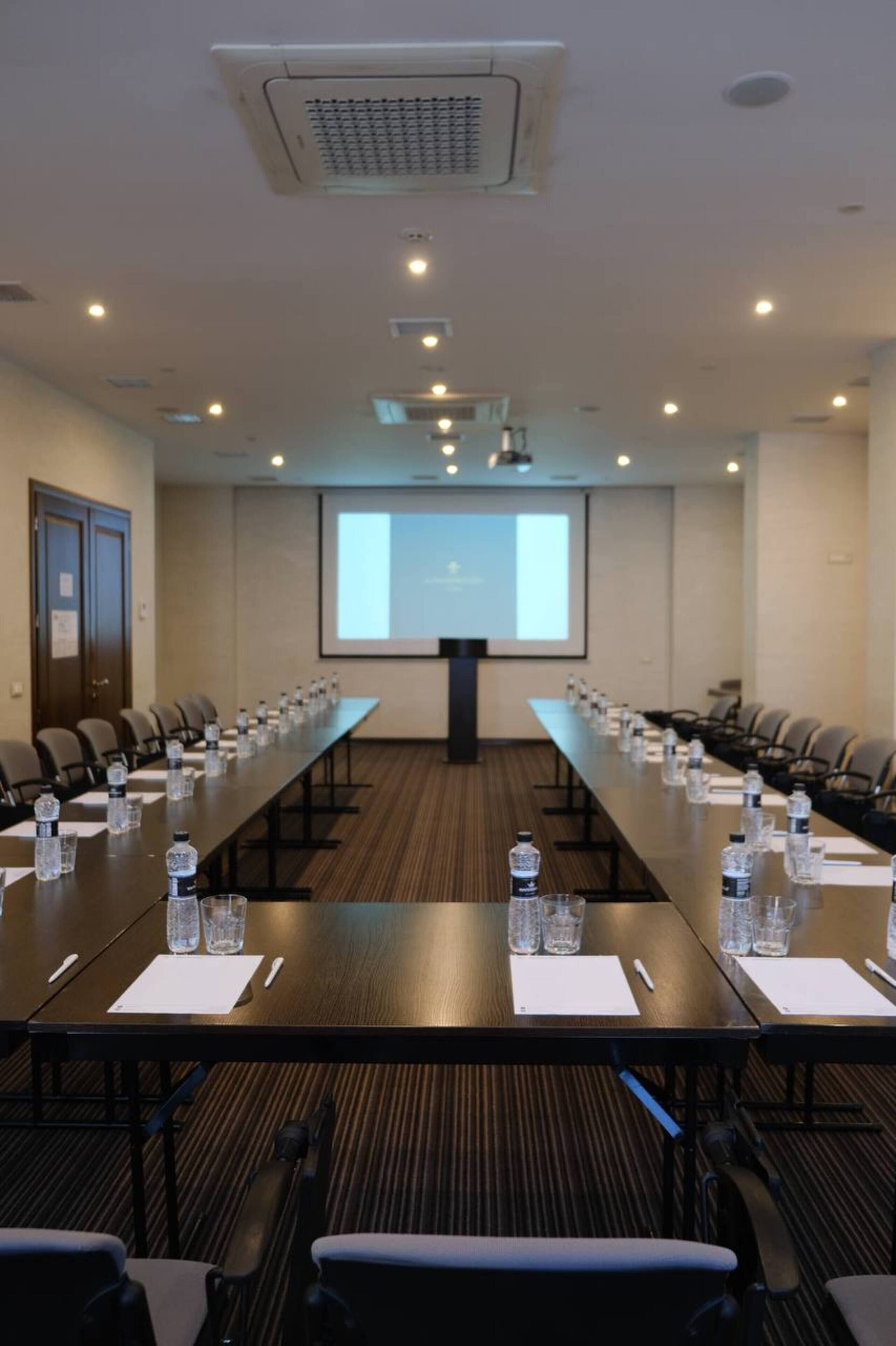 Uma sala de conferência com uma mesa comprida, cadeiras, garrafas de água e uma tela de projetor.