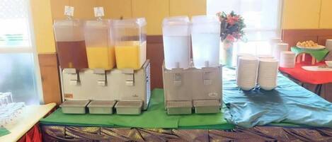 Disposição de buffet com vários distribuidores de bebidas e pilhas de pratos sobre uma mesa.