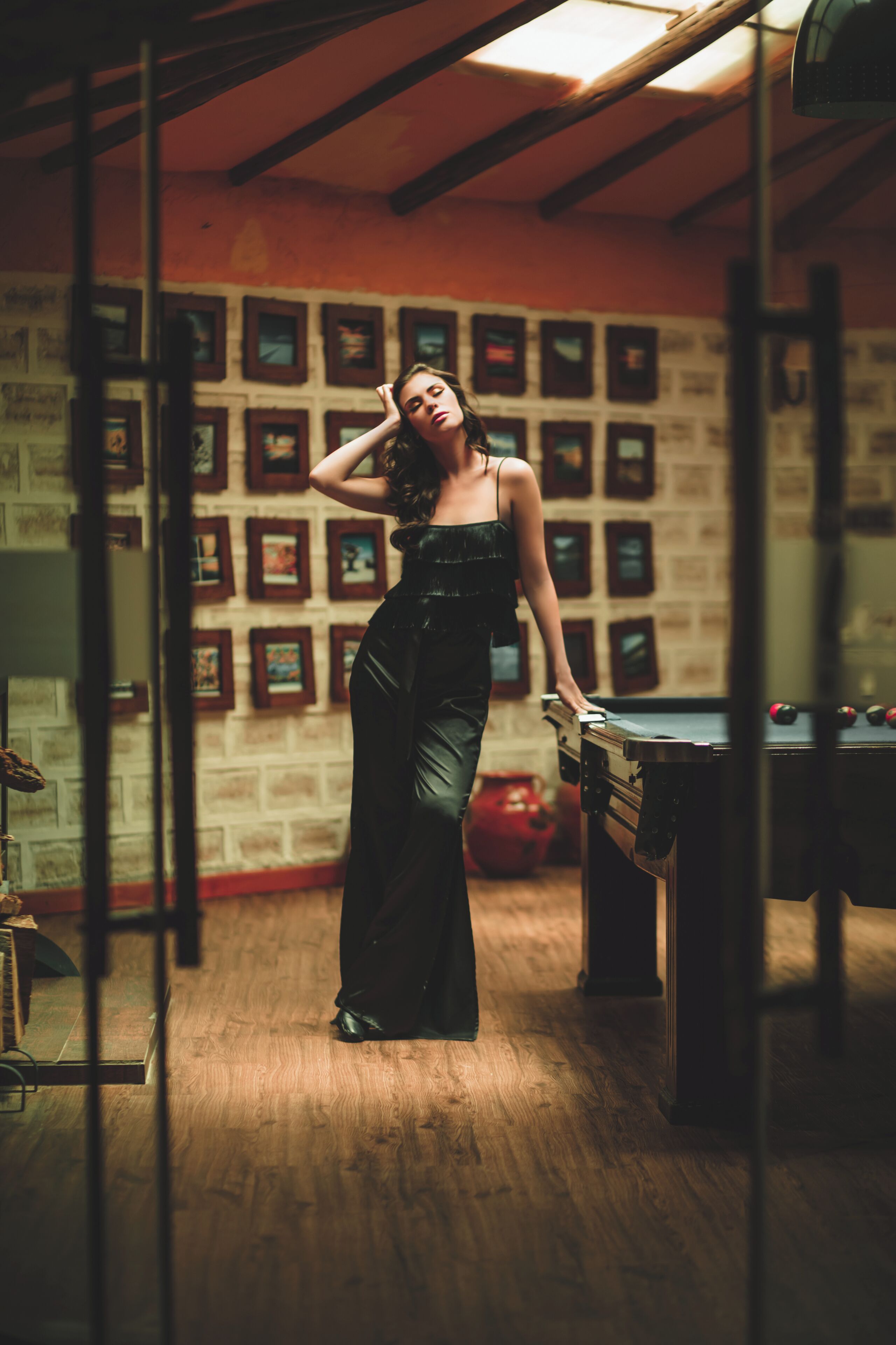 Uma mulher de vestido preto parada em um quarto com mesa de bilhar, piso de madeira e quadros nas paredes.