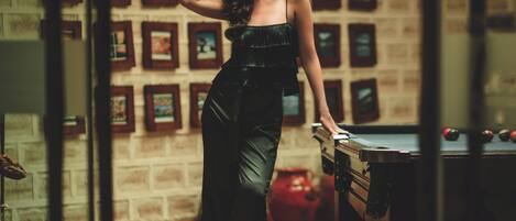 Uma mulher de vestido preto parada em um quarto com mesa de bilhar, piso de madeira e quadros nas paredes.