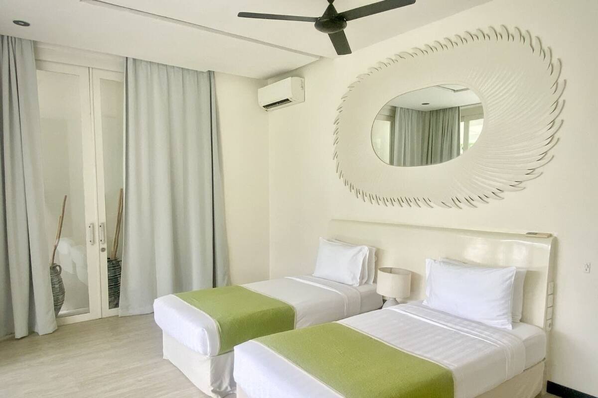 Quarto de hotel com duas camas, um ventilador de teto, um espelho grande e uma janela com cortinas.
