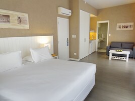 Quarto de hotel com uma cama grande, mesa de cabeceira, sofá, pequena mesa com maçãs, área de cozinha e banheiro visível por uma porta aberta.