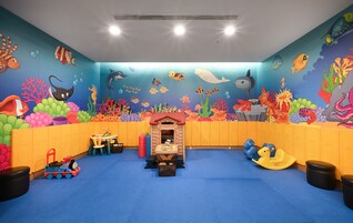 Uma sala de recreação infantil com carpete azul, murais coloridos com vida marinha, uma casinha e diversos brinquedos.