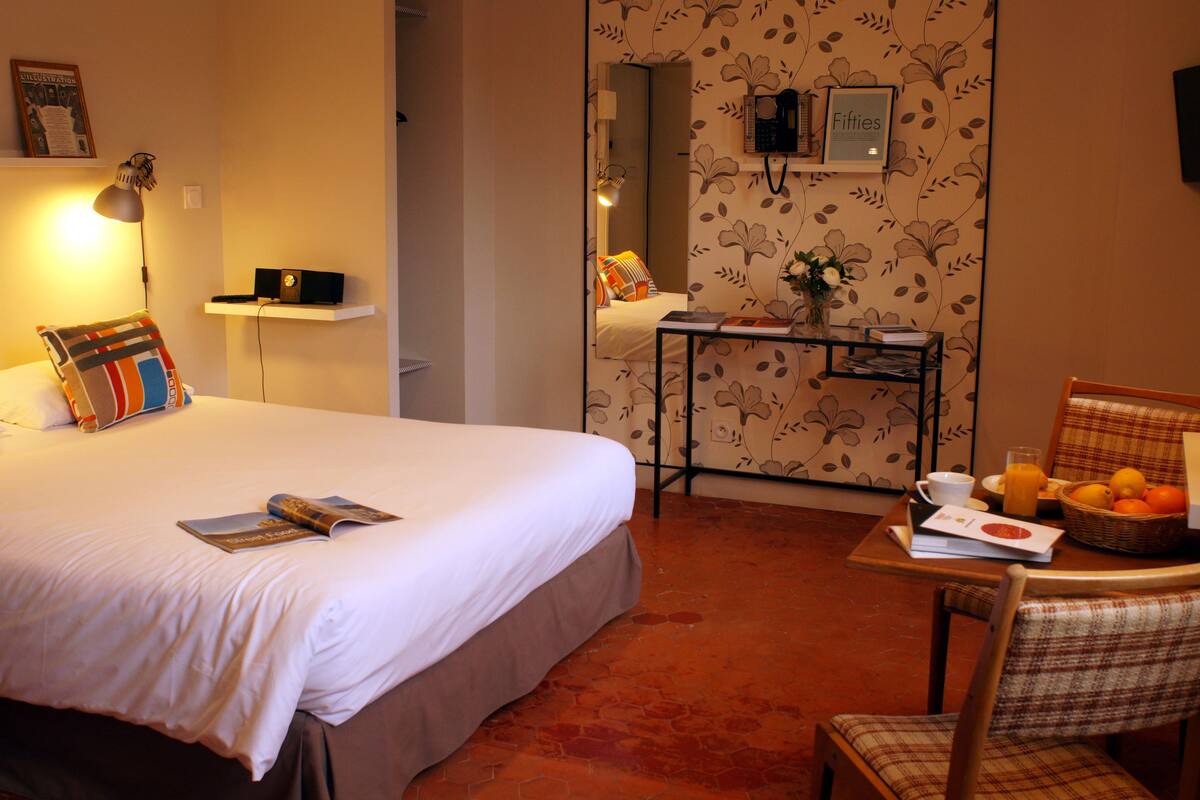 Quarto de hotel com uma cama grande, uma escrivaninha com cadeira, uma pequena mesa com cesta de frutas e um espelho na parede.