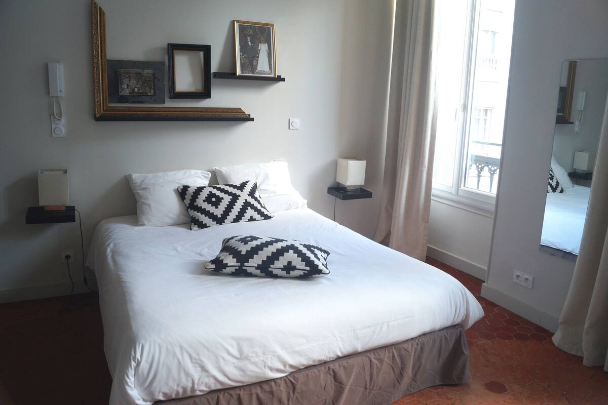 Cama bem arrumada com roupa de cama branca e almofadas estampadas, mesa de cabeceira com abajur e janela com cortinas.