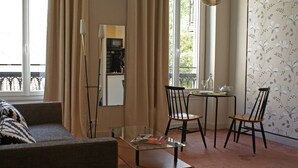 Uma sala de estar com sofá, mesa de centro de vidro, duas cadeiras, uma mesinha com bule e copos, abajur e vista para uma varanda através de grandes janelas.