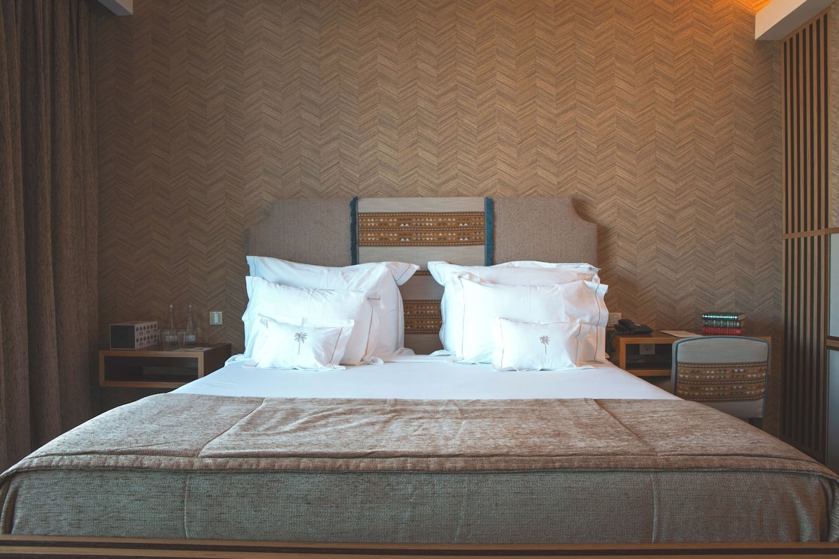 Cama bem arrumada com lençóis e almofadas brancas, cabeceira de madeira com painel decorativo, mesa de cabeceira com abajur e cadeira com enchimento estampado.