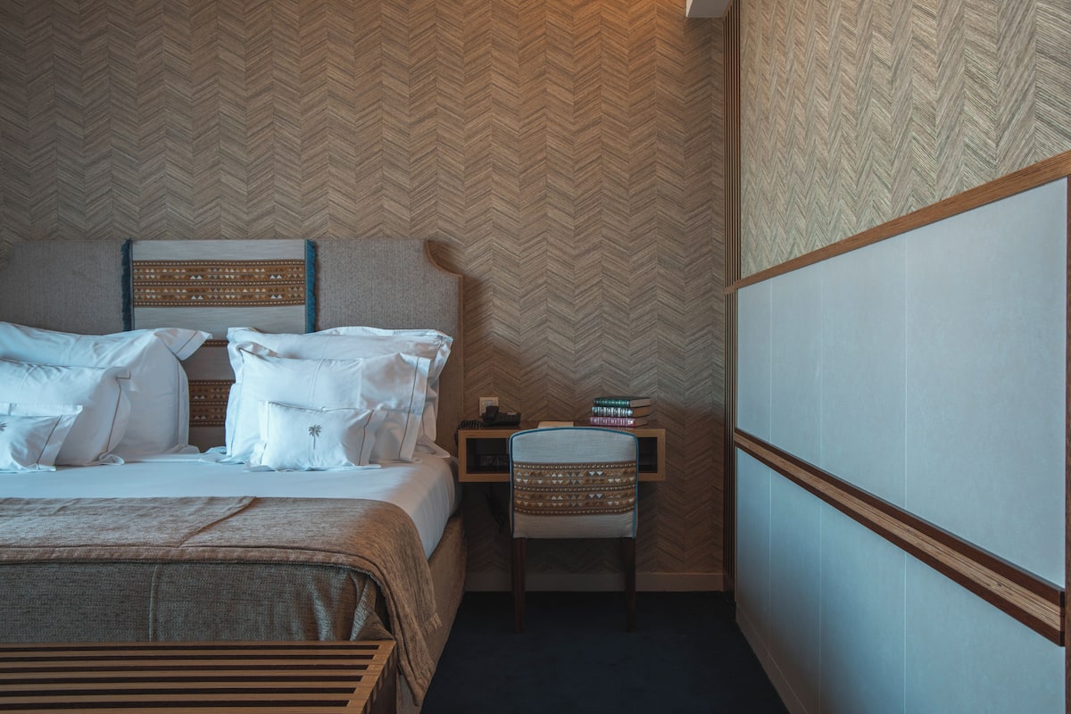 Quarto de hotel com uma cama, uma cadeira, uma mesa de cabeceira e parede com padrão herringbone.