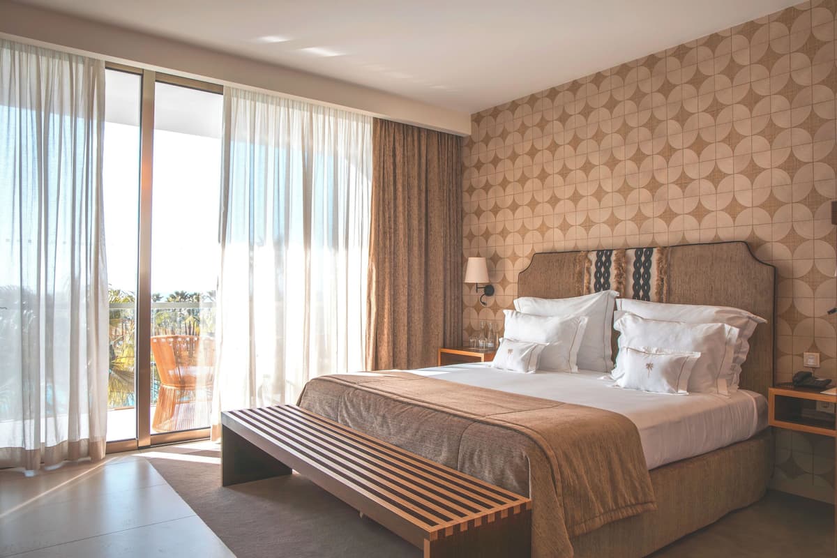 Quarto de hotel moderno com uma cama grande, um banco, uma mesa de cabeceira com abajur e vista para o exterior.