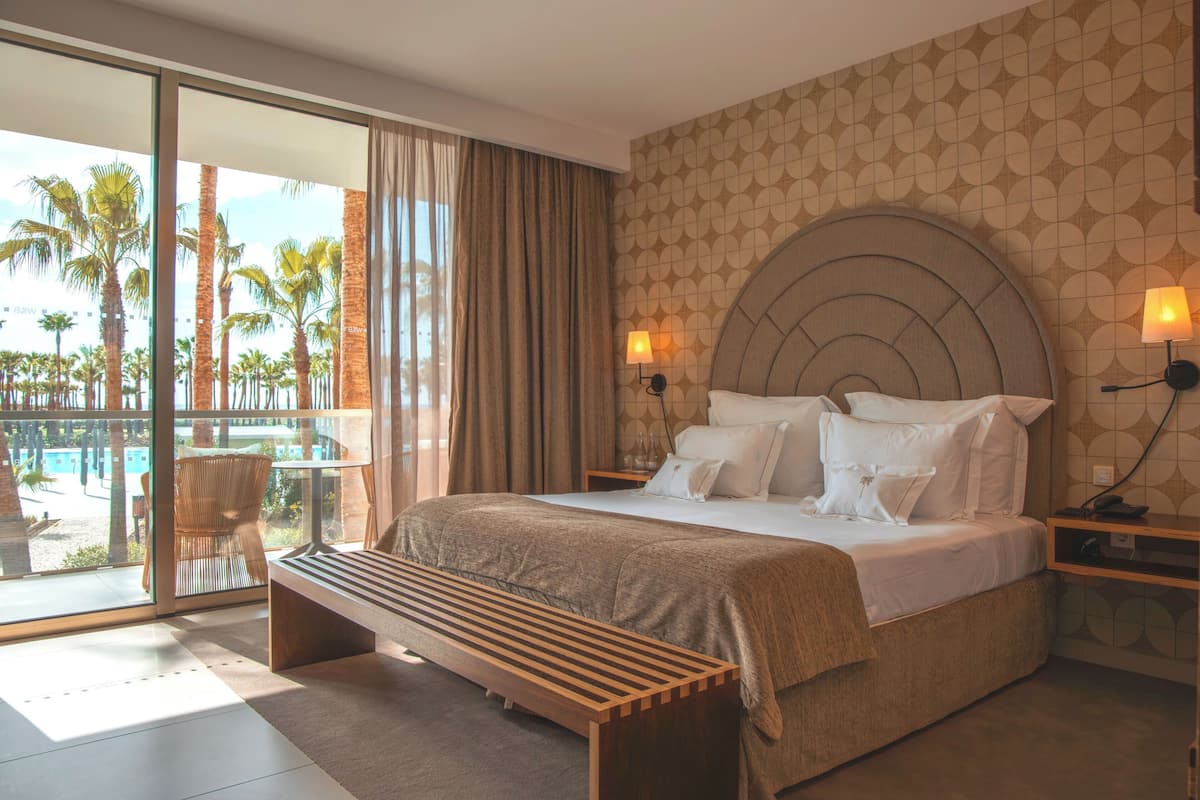 Quarto de hotel com uma cama grande, duas luminárias de cabeceira, um banco e vista para palmeiras e uma piscina lá fora.
