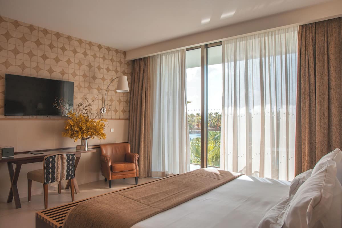Quarto de hotel com cama, escrivaninha com cadeira, televisão na parede e uma janela grande com cortinas.