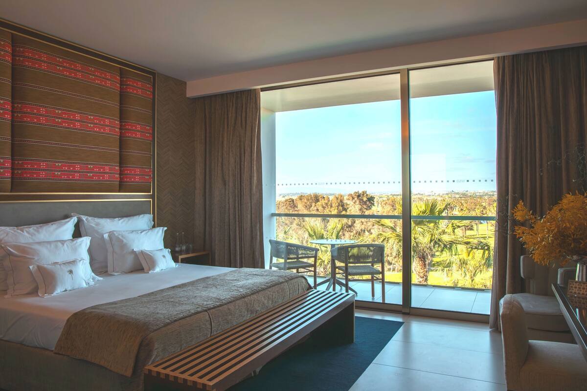 Quarto de hotel com uma cama grande, um banco, uma mesa com cadeiras e varanda com vista para o exterior.