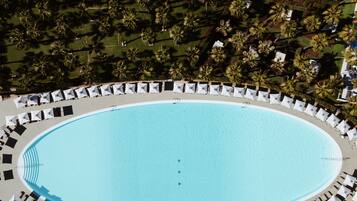 Vista aérea de uma grande piscina oval, rodeada por espreguiçadeiras e guarda-sóis brancos, com palmeiras e um gramado bem cuidado ao fundo.