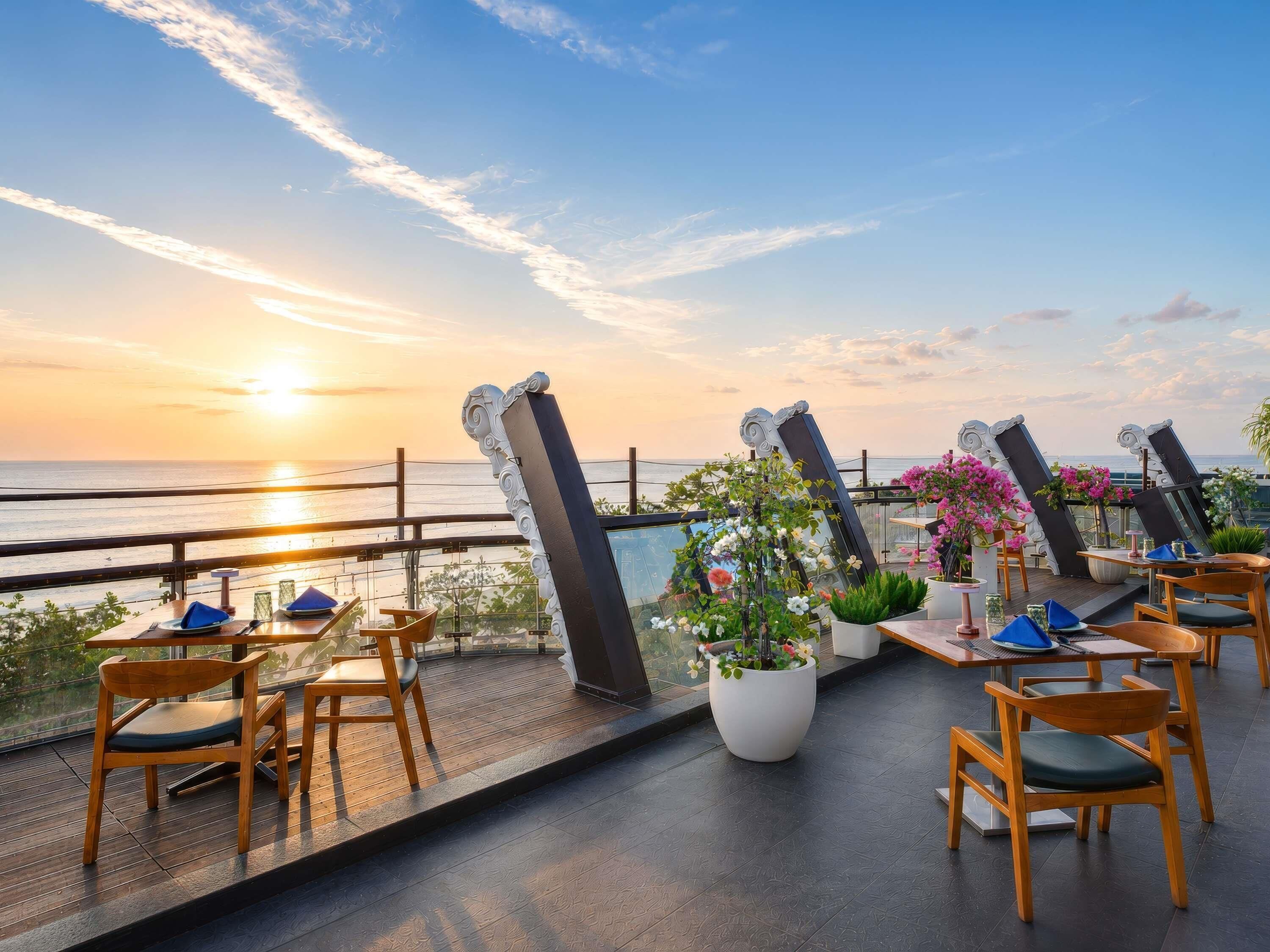 Terraço com mesas e cadeiras, vista para o pôr do sol sobre o mar.
