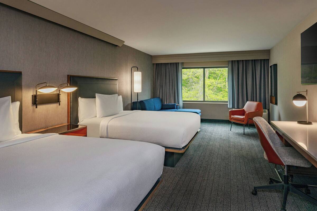 Quarto de hotel com duas camas, uma escrivaninha, uma cadeira e uma janela grande com cortinas.