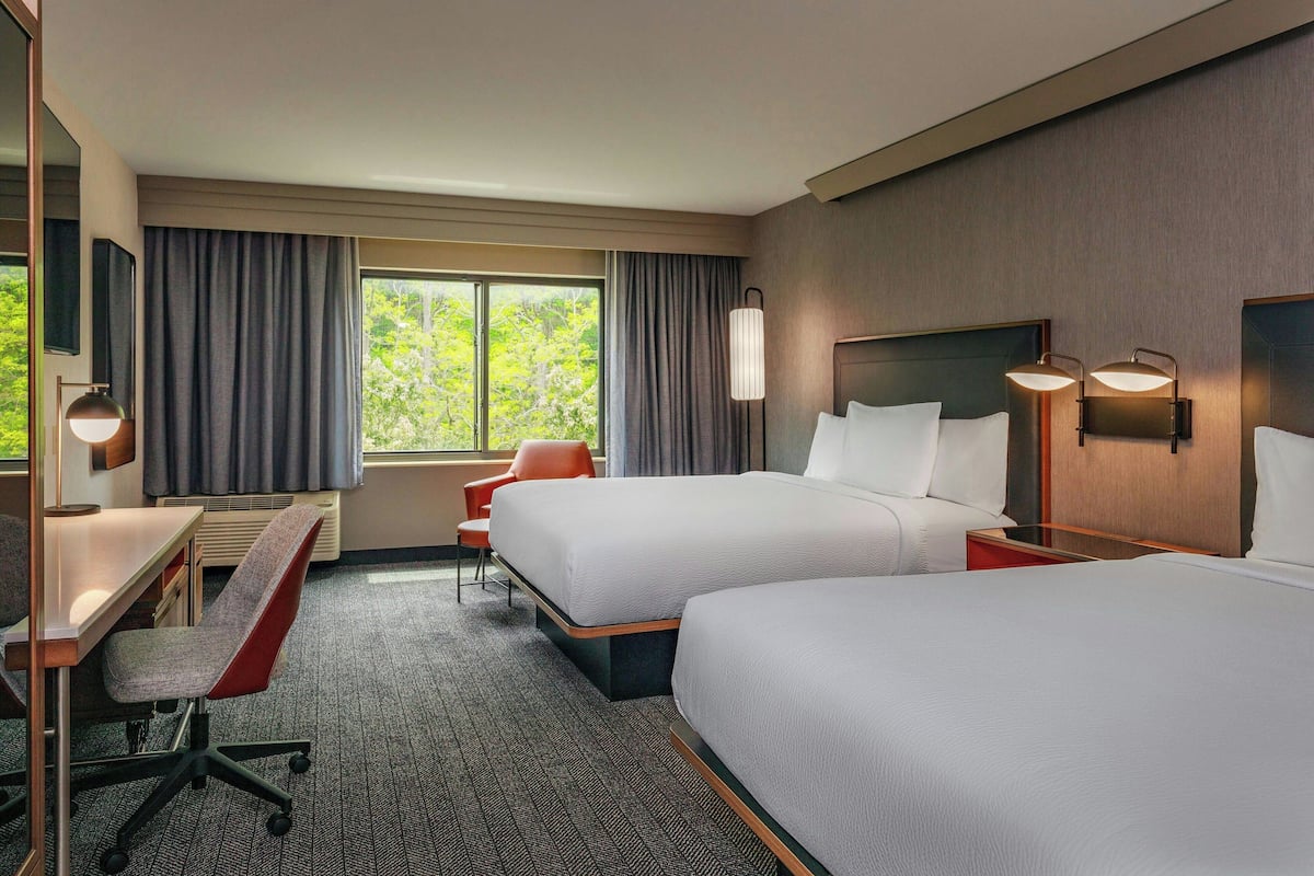 Quarto de hotel com duas camas, uma mesa, uma cadeira e uma janela grande com cortinas.