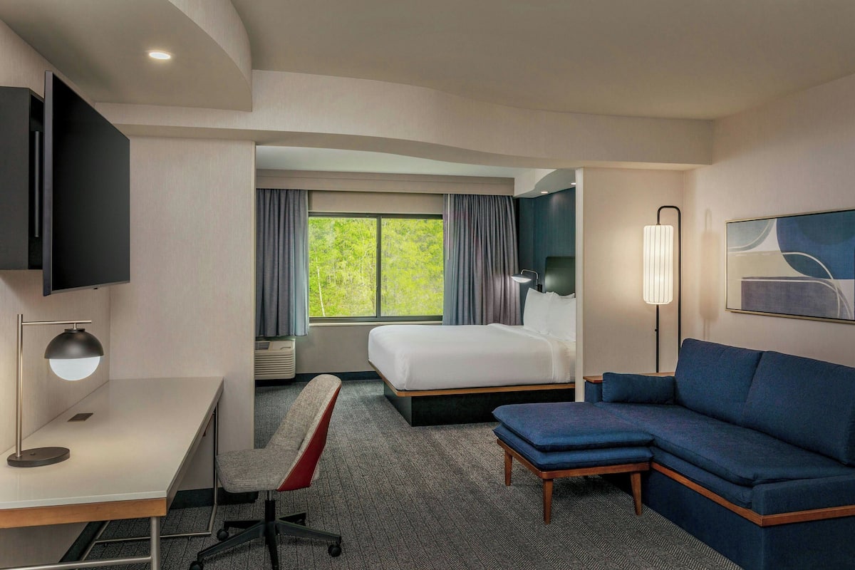 Quarto de hotel com cama, escrivaninha com luminária, cadeira, sofá e televisão na parede.