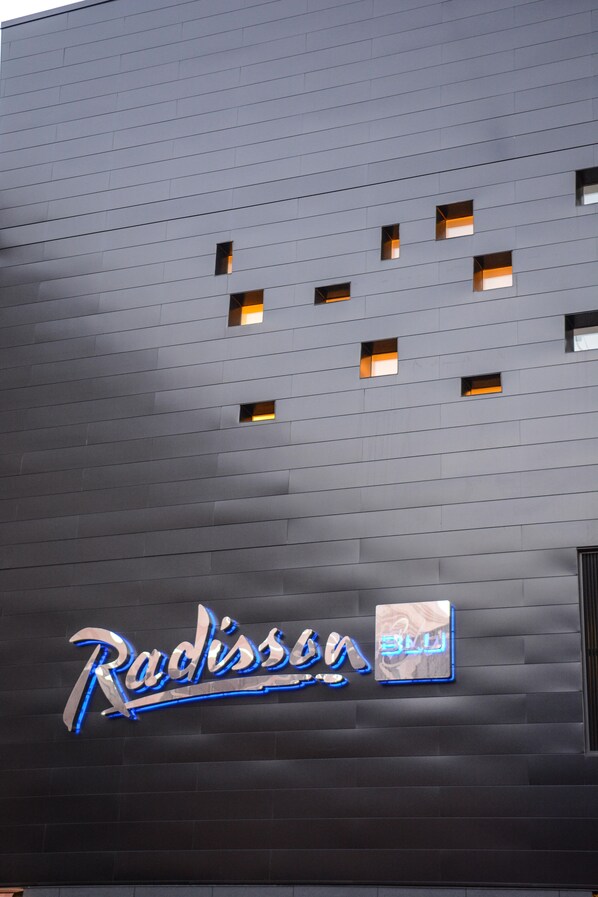 Fachada de edifício com design moderno, apresentando uma série de janelas retangulares uniformemente espaçadas e um letreiro iluminado com o nome "Radisson Blu".