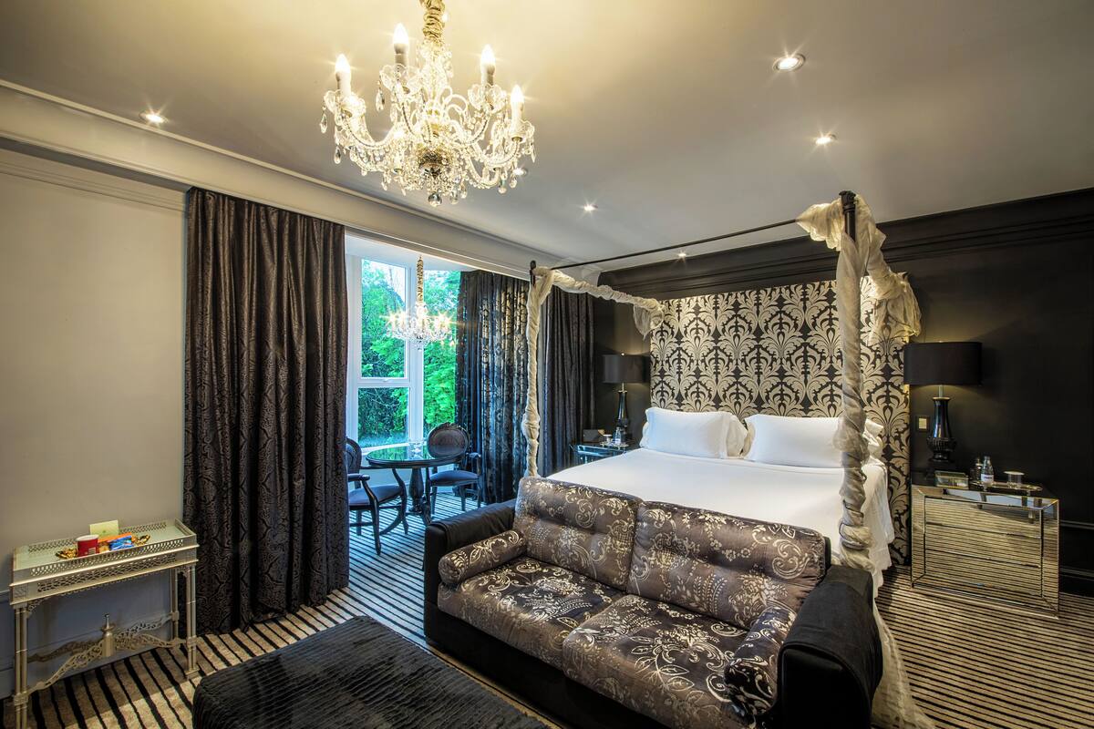 Quarto de hotel com uma cama grande, um sofá, um candelabro, uma mesa com bandeja e vista para o verde lá fora.