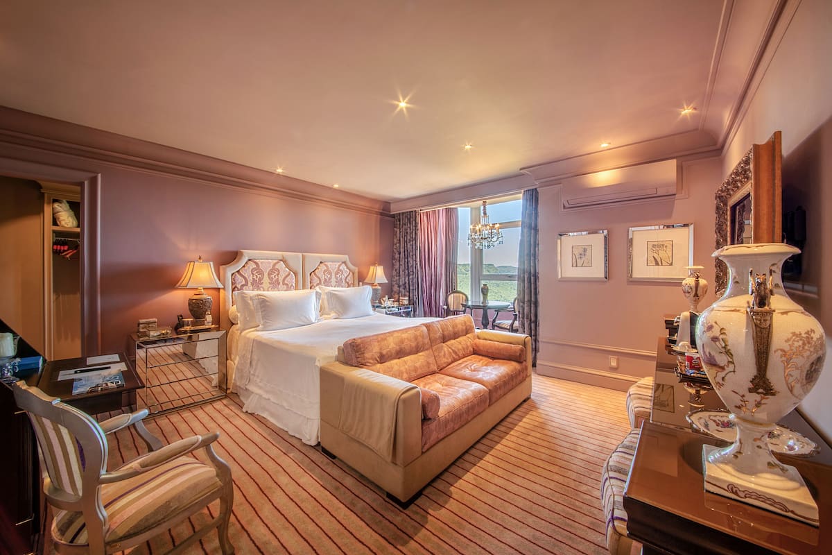 Quarto de hotel com cama, sofá, poltrona, escrivaninha e lustre.