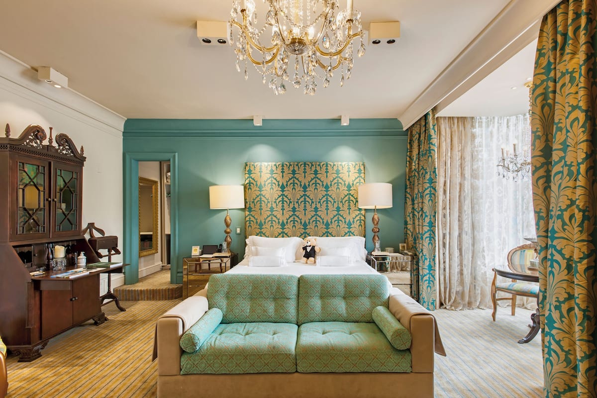 Quarto de hotel com uma cama grande, um sofá verde, uma cômoda de madeira e um lustre.