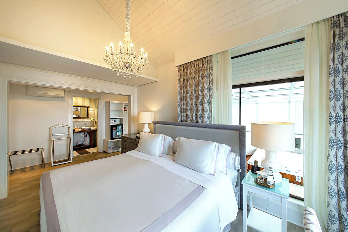 Quarto de hotel com uma cama grande, um candelabro, uma mesa de cabeceira com abajur e vista exterior através de uma janela com cortinas.