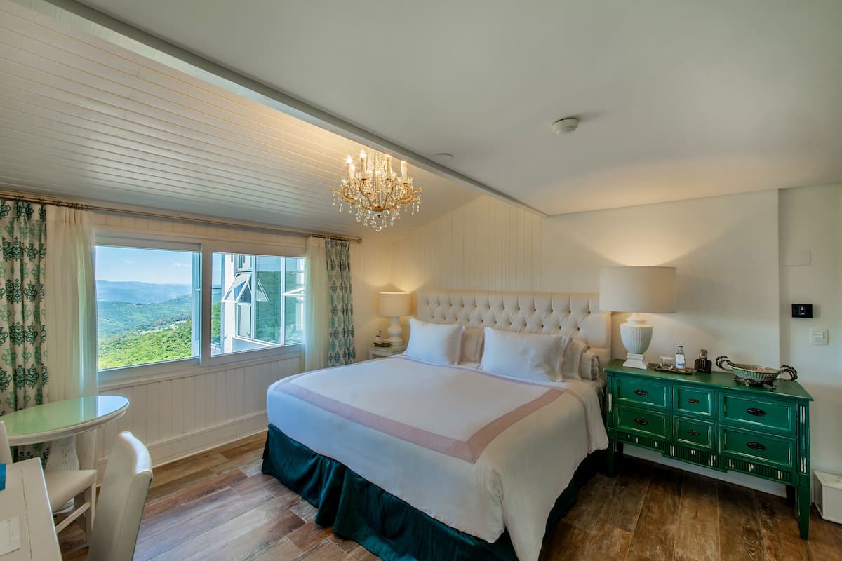 Uma cama grande com cabeceira, um penteoudo verde, uma mesa com cadeiras e vista para o exterior pela janela.
