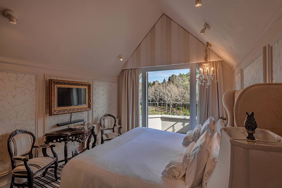 Quarto de hotel com uma cama grande, televisão, escrivaninha e vista para árvores pela janela.