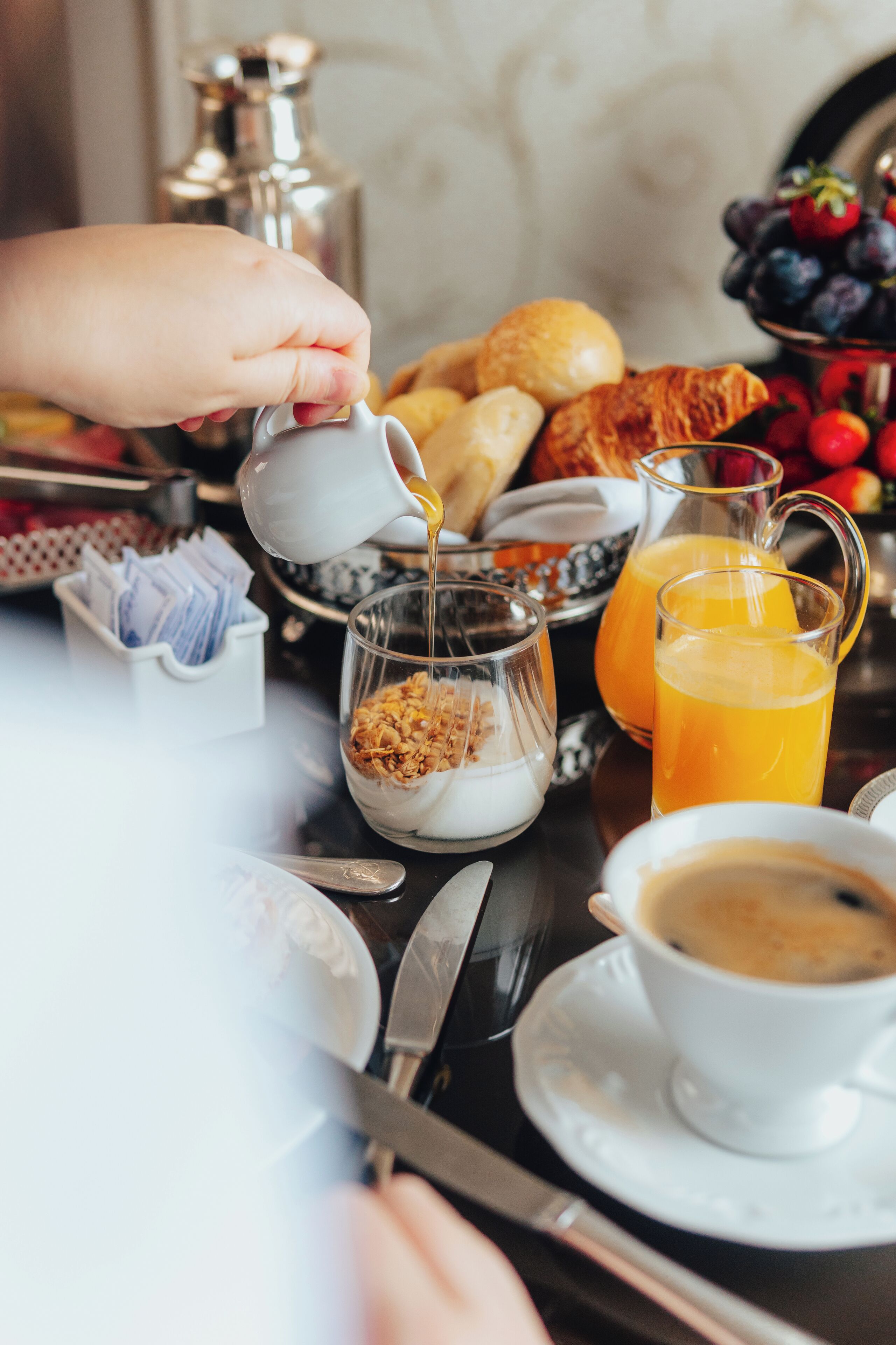 Mesa de café da manhã com uma mão servindo leite em um copo, jarra de suco de laranja e diversas opções de pães e frutas na mesa.