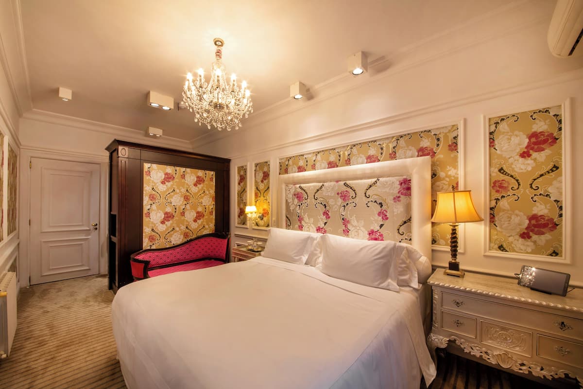 Quarto de hotel com cama grande, lustre, papel de parede floral e mesa de cabeceira com luminária.