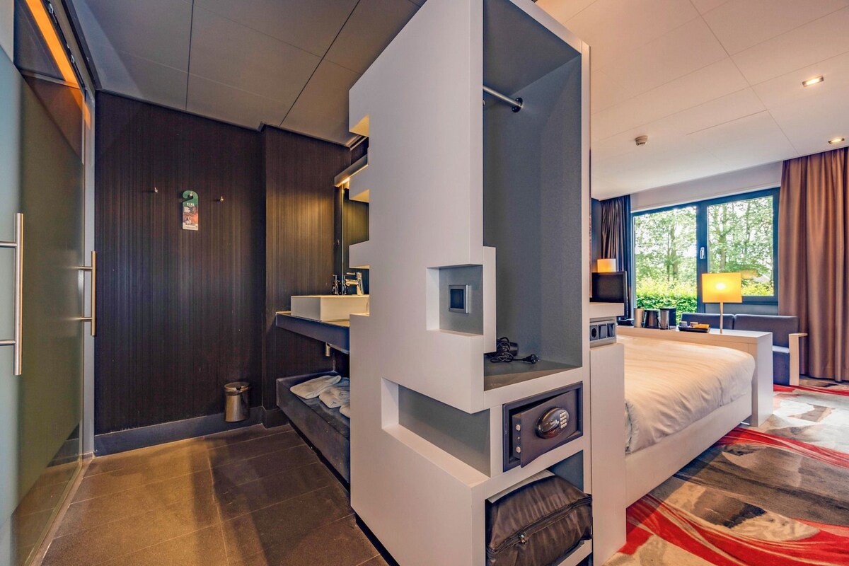 Quarto de hotel moderno com cama, escrivaninha, toucador e área de chuveiro.