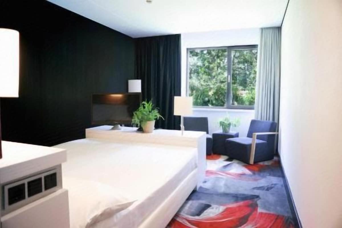 Quarto de hotel com cama, escrivaninha, cadeira, janela e planta.